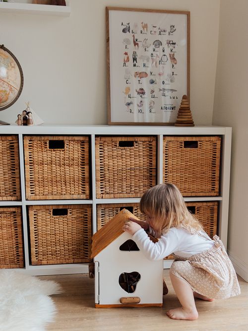 Mädchen vor einem Regal mit Körben das mit einem Puppenhaus spielt.
