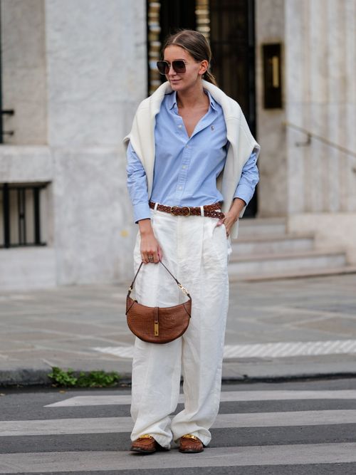 Diane Batoukina trägt eine blaue Bluse, einen weißen Pulli über die Schultern, eine weiße Bundfaltenhose, braunen Gürtel, braune Handtasche aus Wildleder, braune Loafer und eine Pilotensonnenbrille in Paris.