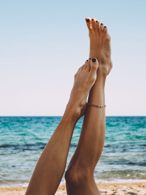 Frauenbeine mit Fußkettchen, die am Strand ins Meer ragen.