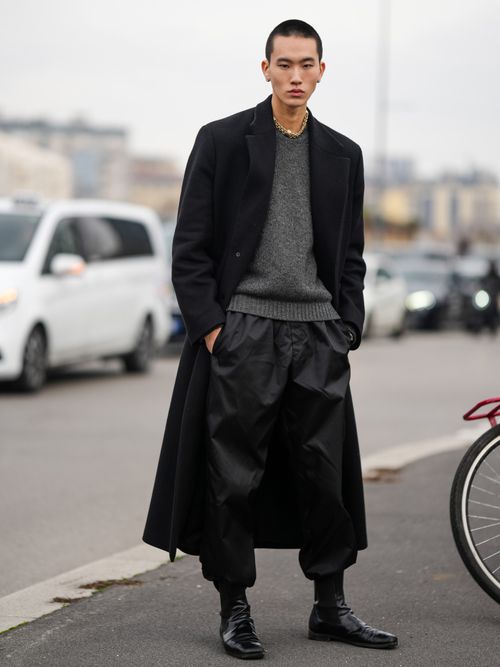 Street Style Look von der Milan Fashion Week, wo ein Mann einen schwarzen Mantel, grauen Pulli, schwarze Cargohose und schwarze flache Boots trägt.