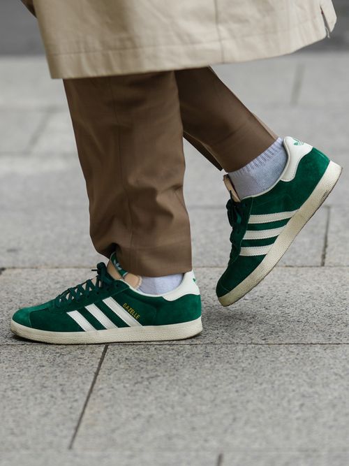 Close-up von einem Mann der eine braune Hose, beigen Trenchcoat, weiße Socken und Adidas Gazelle Sneaker trägt.