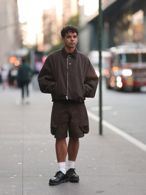 Street Style Look von der New York Fashion Week, wo ein Gast eine braune Bomberjacke, braune Cargoshorts, weiße Socken und schwarze Lederschuhe trägt.