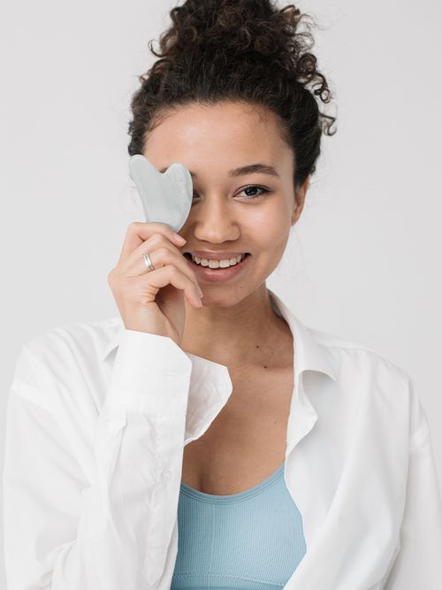Frau in weißer Bluse mit blauem Top hält einen Gua Sha Stein vors Auge.