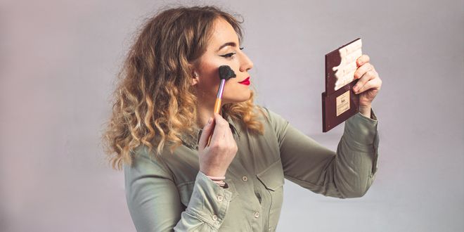 Frau in grünem Oberteil hält eine Schminkpalette vor sich und schminkt sich mit einem Pinsel die Wangen.