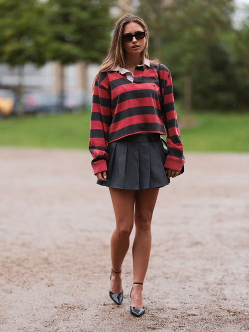 Street Style Look von der Copenhagen Fashion Week, wo ein Gast ein gestreiftes Langarmshirt, Faltenrock, Sonnenbrille und Lacklederpumps trägt.