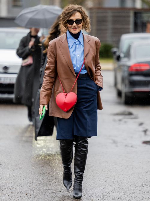 Street Style Look von einem Fashion Week Gast, der eine braunen Lederblazer, blaue Bluse und dunkelblaue Bermudashorts mit schwarzen Lederstiefel und einer herzförmigen Alaia Tasche in Copenhagen trägt.