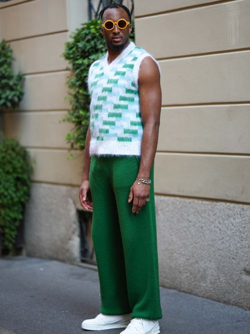 Street Style Look von der Milan Fashion Week, wo ein Gast einen karierten Pullunder, grüne Hose, weiße Sneaker und orangefarbene Sonnenbrille trägt.