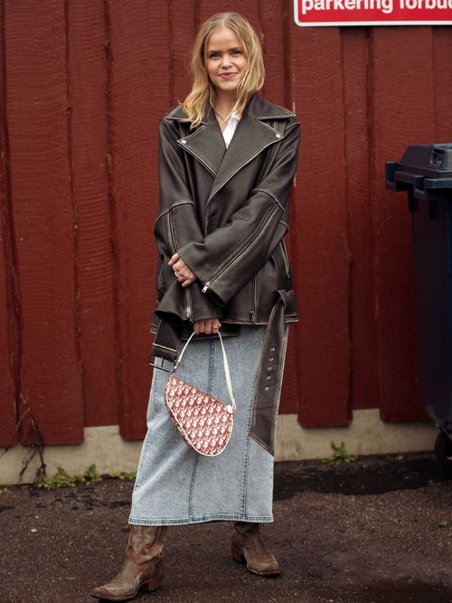 Street Style Look von der Copenhagen Fashion Week, wo ein Gast eine Lederjacke, mit langem Jeansrock, Cowboy Boots und einer Dior Tasche trägt.