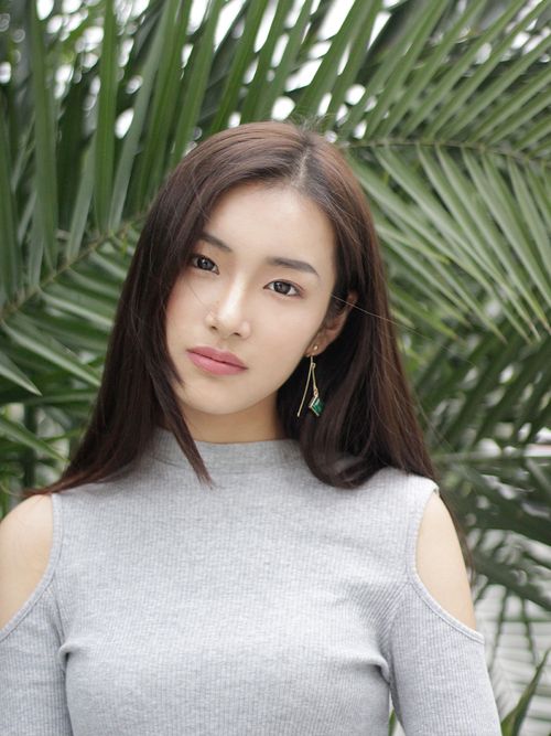 Frau mit braunen offenen Haaren und einem grauen Shirt mit Cut-outs steht vor einer Palmenpflanze.